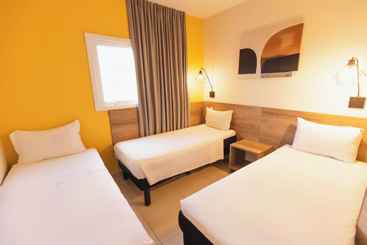 Quarto de hotel com três camas, janela com cortinas, luminária na parede e uma mesa pequena.