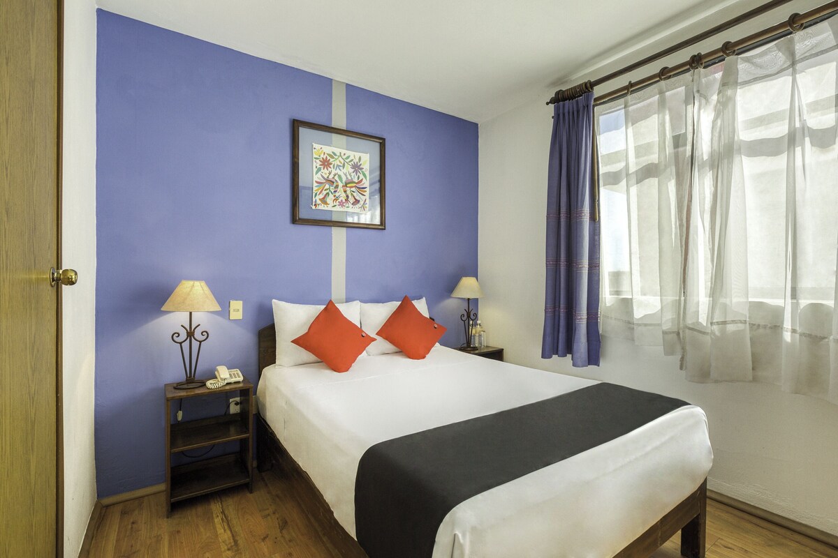 Quarto de hotel com cama, duas mesinhas de cabeceira, um criado-mudo, um quadro na parede e janela com cortinas.