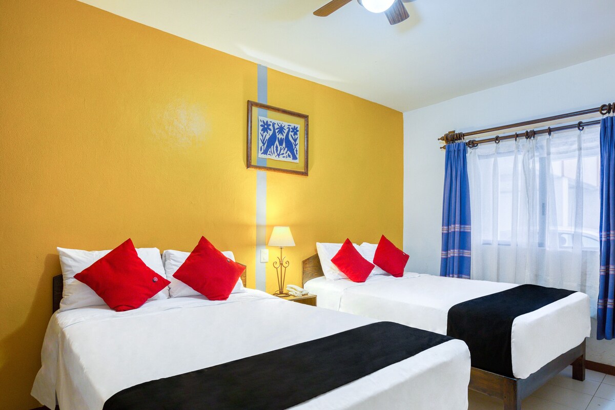Quarto de hotel com duas camas, cada uma com roupa de cama branca e almofadas vermelhas, um ventilador de teto, uma mesa de cabeceira com luminária e uma janela com cortinas azuis.