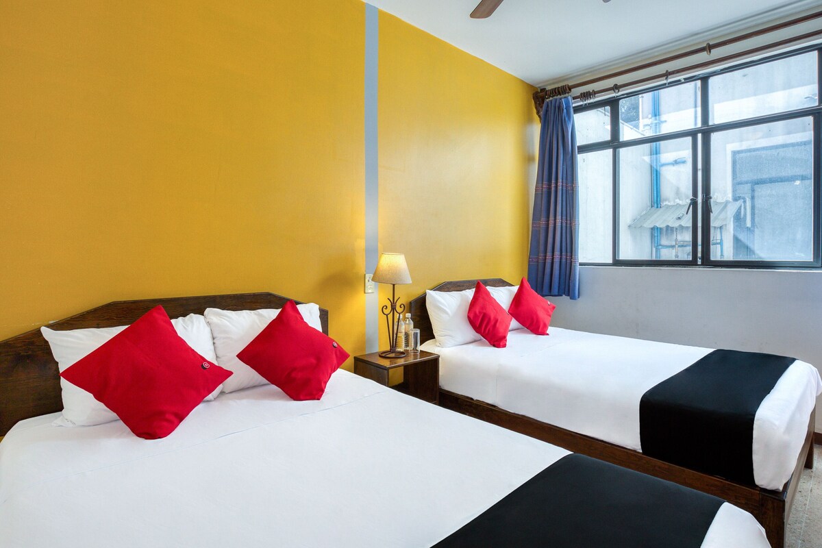 Quarto de hotel com duas camas, um ventilador de teto, uma mesa de cabeceira com abajur e uma janela com cortinas.