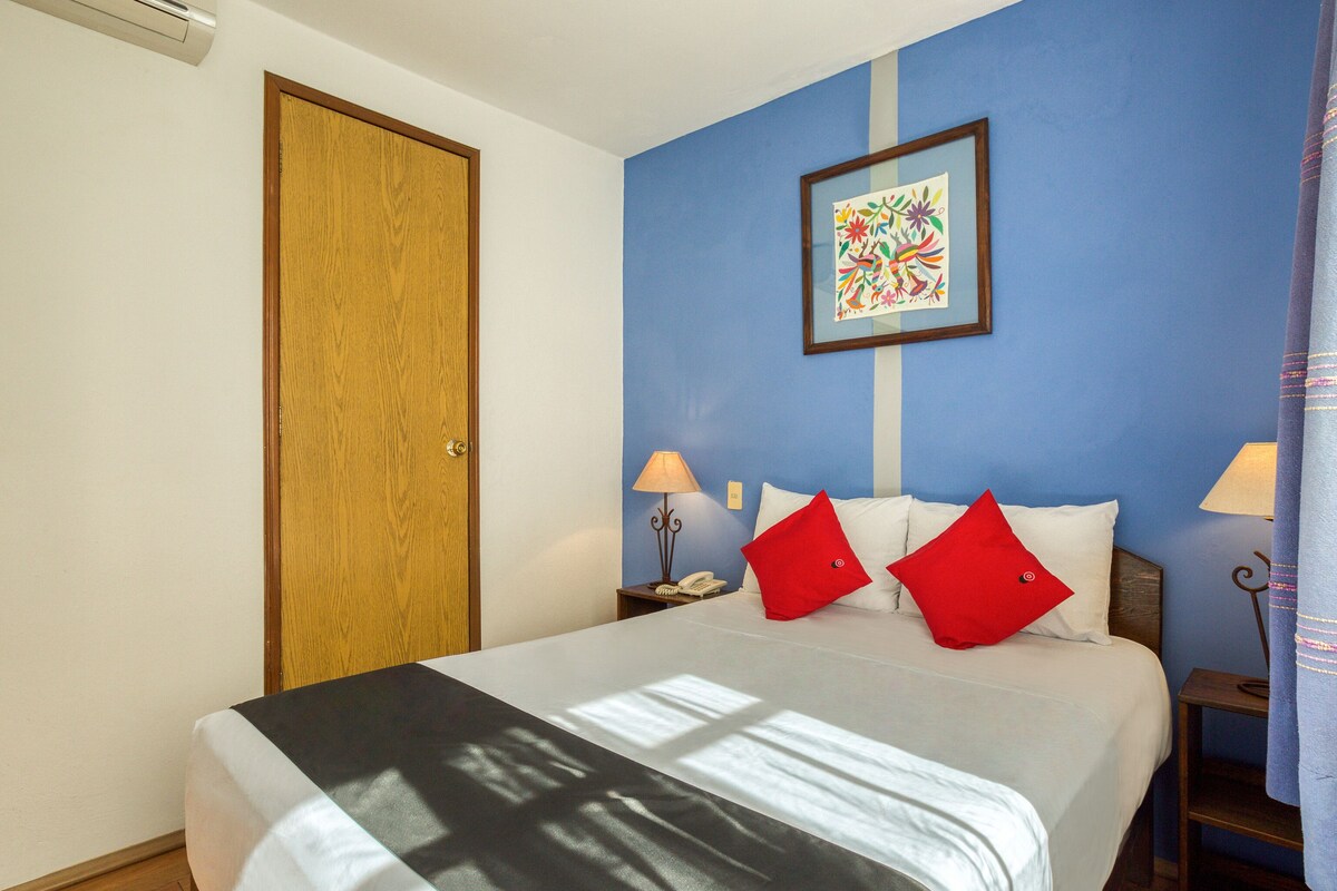 Quarto de hotel com cama, porta de madeira, parede azul, quadro e mesa de cabeceira com abajur.