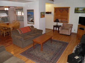 Uma sala de estar com sofá, poltrona, mesa de centro e televisão. Há também um espaço de jantar com mesa e cadeiras.
