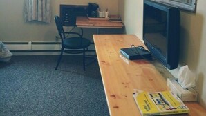 Um quarto pequeno e pouco mobiliado, com uma mesa, uma cadeira, uma televisão e uma janela com cortinas.