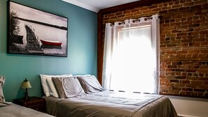 Quarto com cama, mesa de cabeceira, luminária, pintura e parede de tijolos.