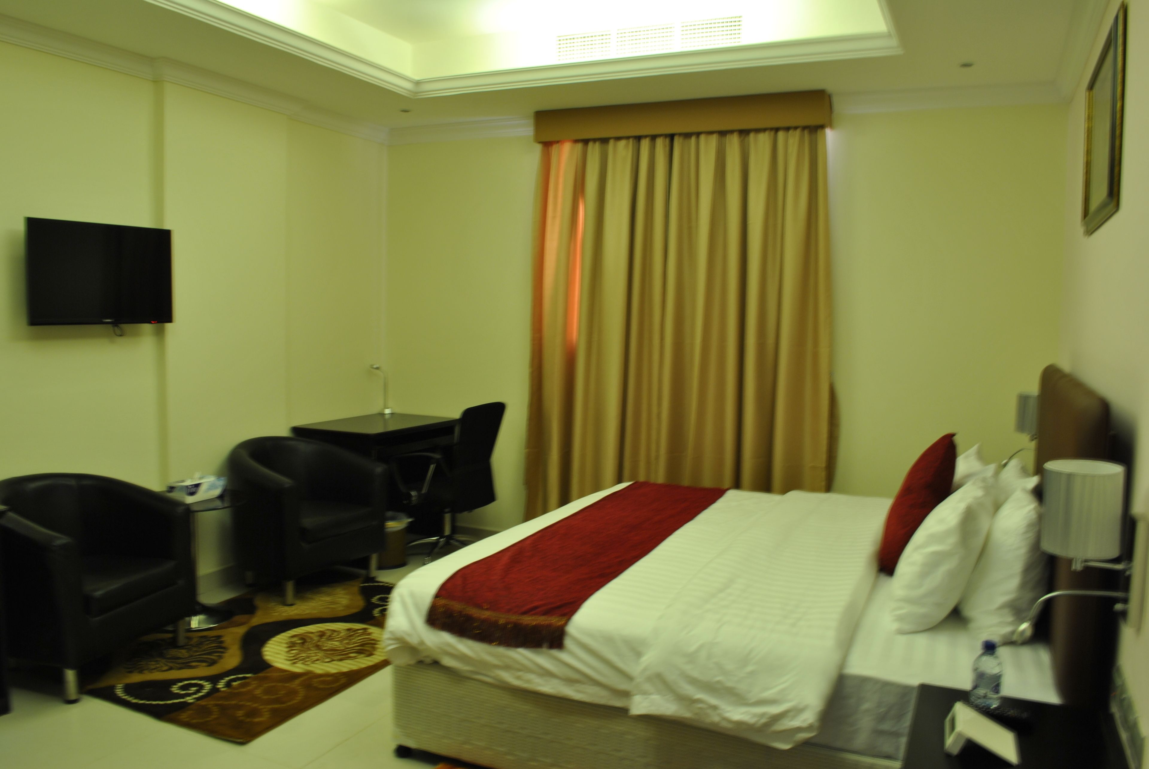 Quarto de hotel com cama, escrivaninha, duas cadeiras, televisão e janela com cortinas.