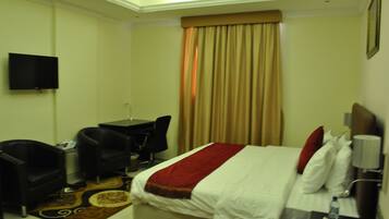 Quarto de hotel com cama, escrivaninha, duas cadeiras, televisão e janela com cortinas.