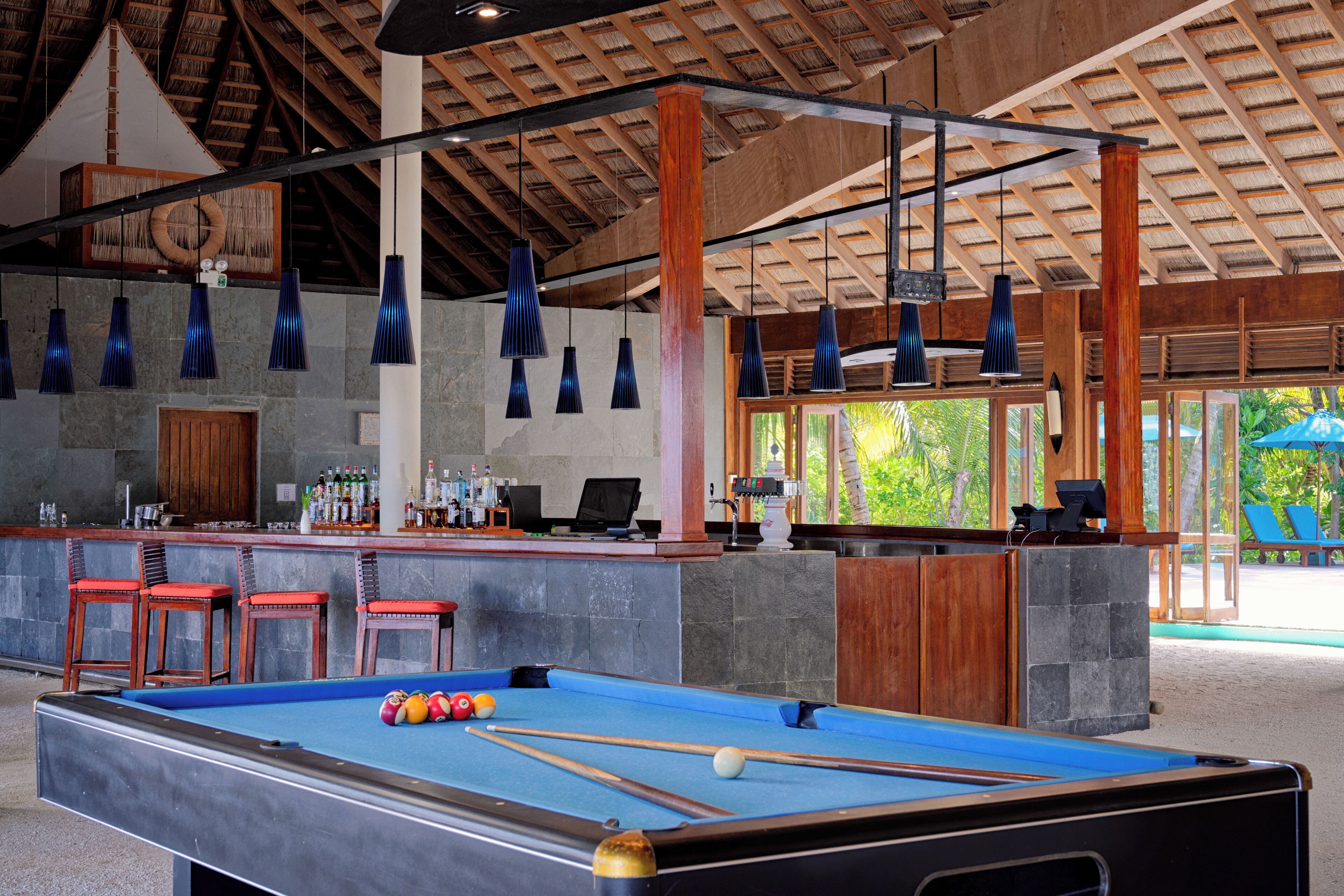 Uma mesa de bilhar com bolas de bilhar, área de bar com banquetas e luminárias pendentes azuis.