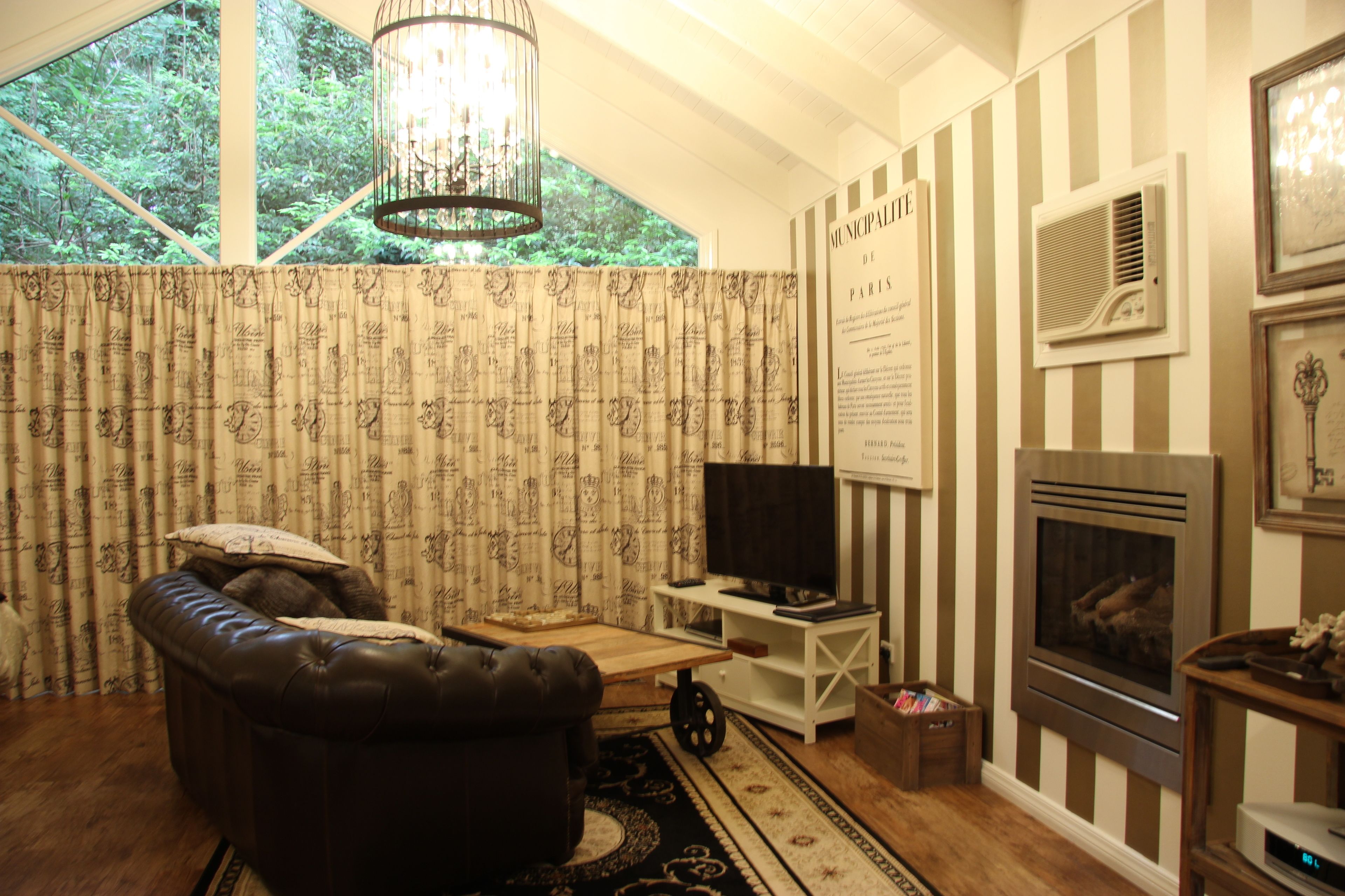 Uma sala de estar com um sofá de couro, uma televisão sobre um suporte branco, uma lareira e uma janela grande com cortinas.