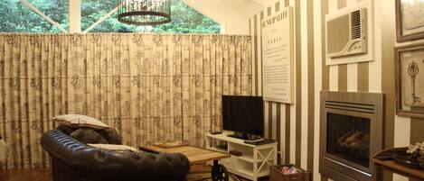Uma sala de estar com um sofá de couro, uma televisão sobre um suporte branco, uma lareira e uma janela grande com cortinas.