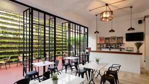 Uma área de refeições interna com mesas e cadeiras, um balcão de cozinha com diversos objetos, e uma grande janela com vista para o verde.