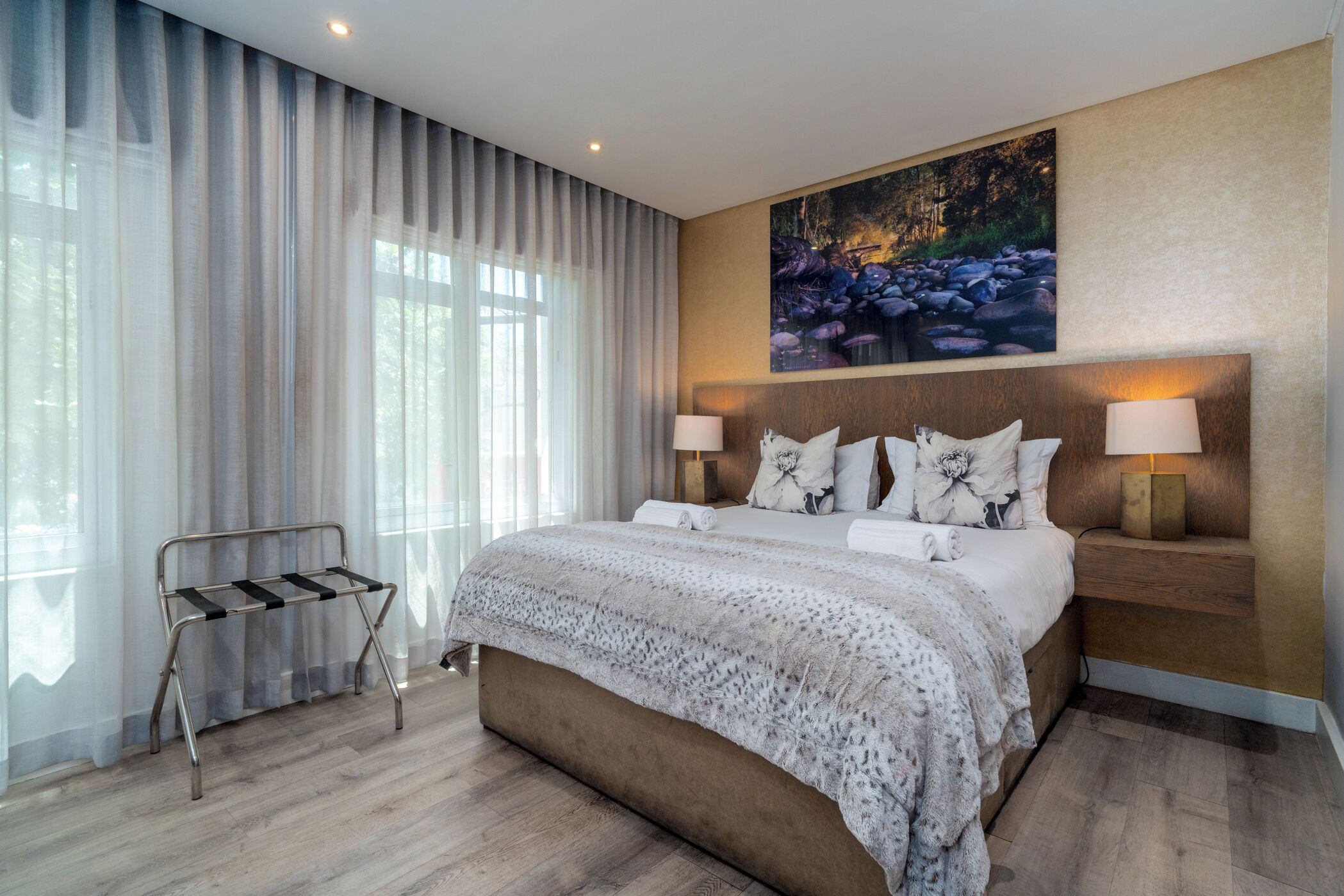 Quarto com uma cama grande, duas luminárias de cabeceira, um banquinho e um quadro na parede.