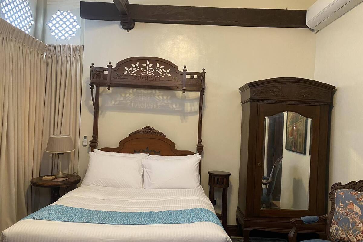 Cama bem arrumada com cabeceira de madeira, abajur de apoio e pequena mesa com luminária. Também se vê um espelho grande e uma cadeira.