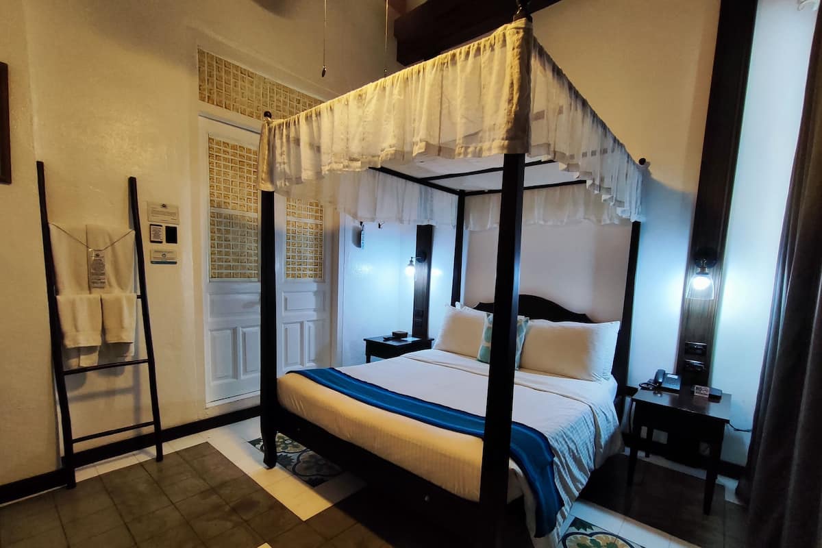 Uma cama de dossel com cortina, uma mesa de cabeceira com abajur, um telefone fixado na parede e um porta-toalhas.