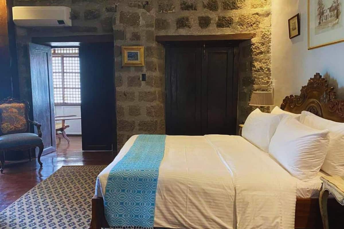 Quarto com cama, uma cadeira, guarda-roupa de madeira e parede de pedra.