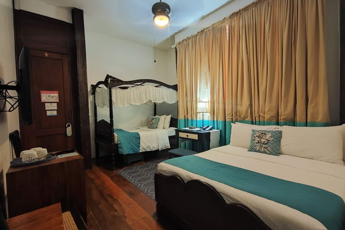 Quarto de hotel com uma cama de dossel, uma cama menor, uma escrivaninha com abajur e piso de madeira.