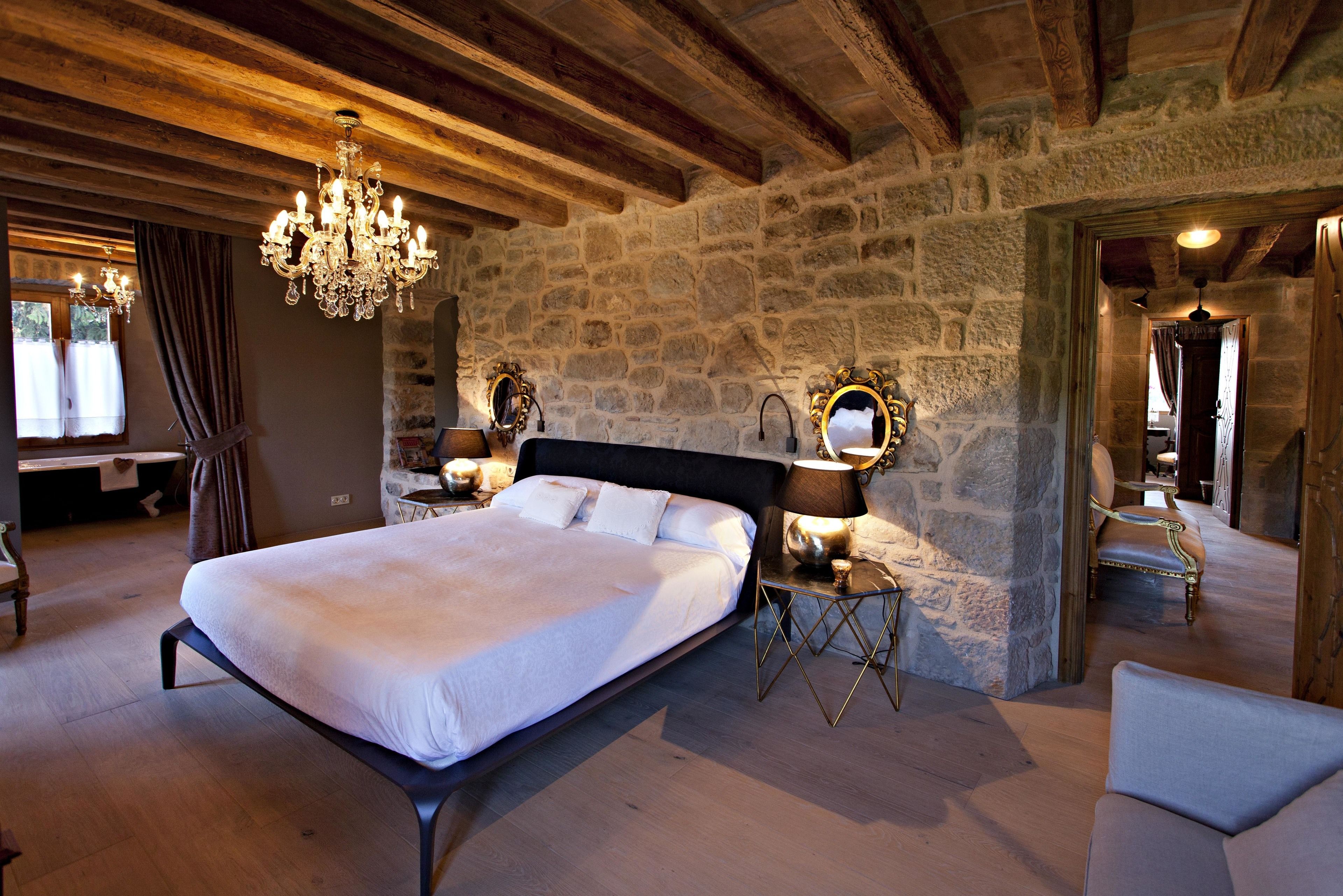 Um quarto espaçoso com uma cama grande, paredes de pedra, vigas de madeira, um candelabro e um espelho.