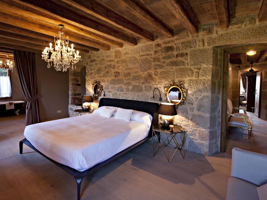 Um quarto espaçoso com uma cama grande, paredes de pedra, vigas de madeira, um candelabro e um espelho.