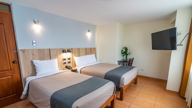 Quarto de hotel com duas camas, uma cabeceira de madeira, televisão de tela plana, uma mesinha com uma planta e uma porta.