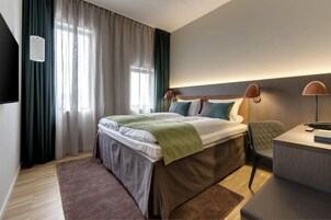 Quarto de hotel com cama, escrivaninha, cadeira e televisão.