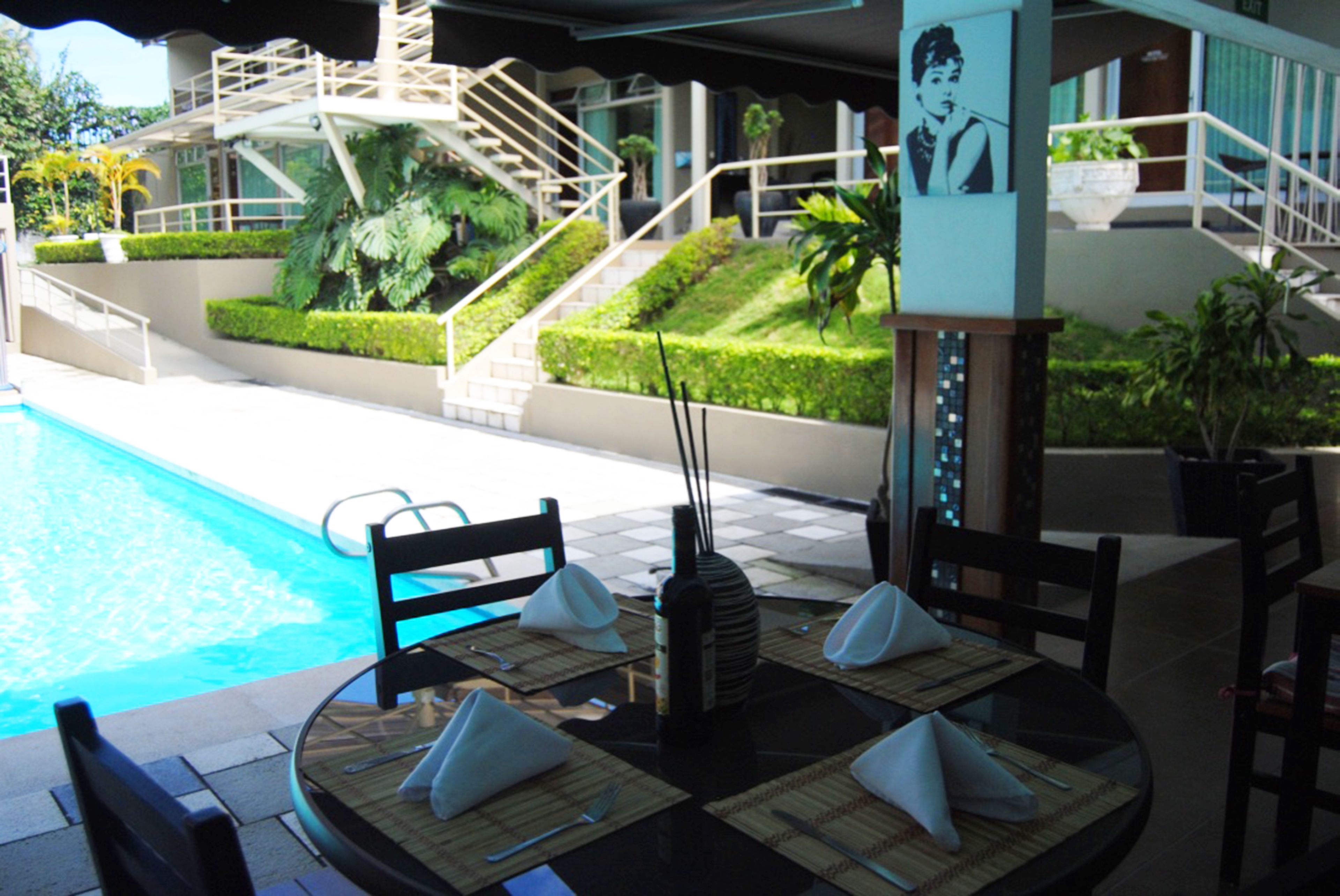 Uma área de refeições com mesas preparadas para dois, perto de uma piscina, com uma escadaria e vegetação ao fundo.