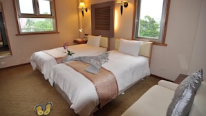 Quarto de hotel com duas camas, um sofá, uma janela, uma mesa de cabeceira e um par de chinelos no chão.