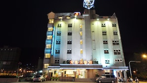 Um hotel com vários andares, com uma luminária geométrica proeminente no telhado, janelas iluminadas e uma área de entrada bem iluminada.