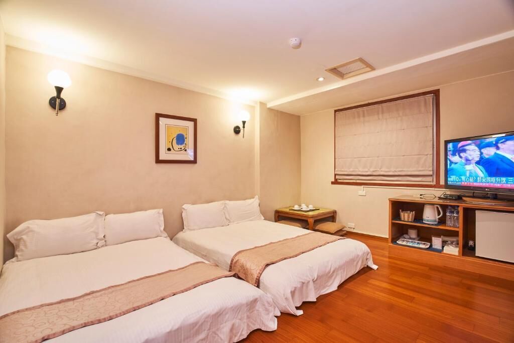 Quarto de hotel com duas camas, televisão, uma mesa pequena com abajur e janela com persianas.