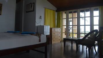 Quarto com cama, cómoda de madeira, escrivaninha e uma cadeira. Há uma janela com cortinas amarelas e vista para o exterior.