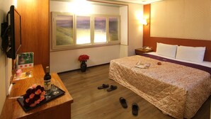 Quarto de hotel com cama, escrivaninha com um vaso de flores, janela com vista e chinelos no chão.