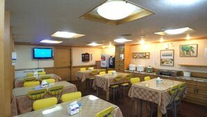 Uma sala de jantar com várias mesas e cadeiras, uma televisão presa na parede e eletrodomésticos visíveis ao fundo.
