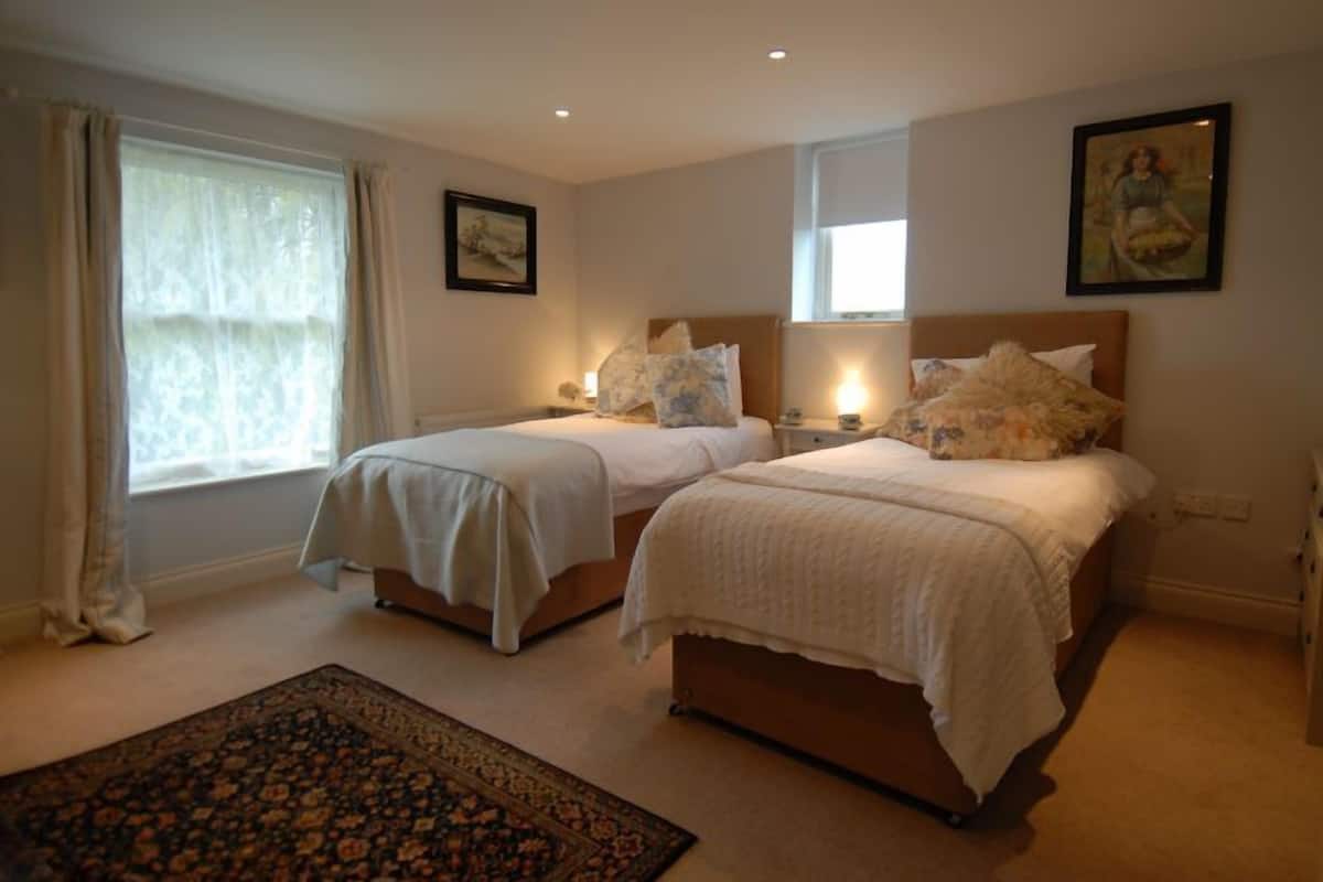 Quarto de hotel com duas camas, uma janela grande com cortinas, um tapete e obras de arte nas paredes.