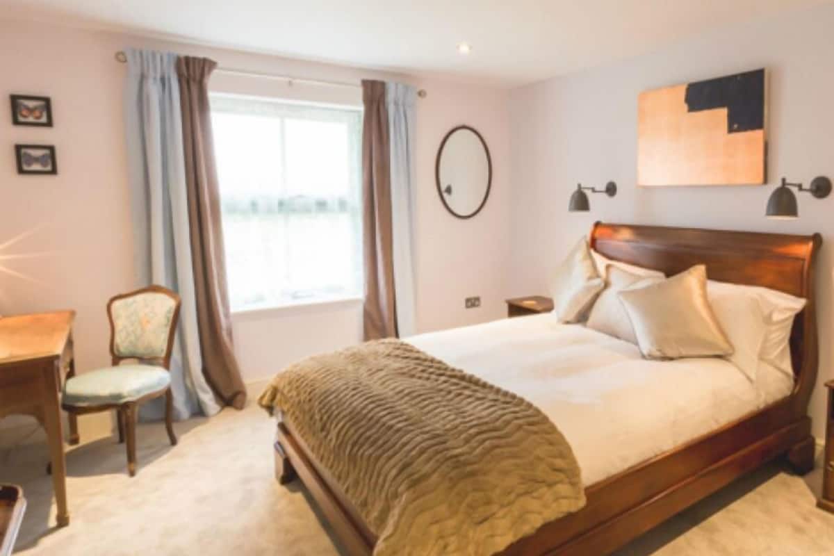 Quarto de hotel com uma cama grande, uma escrivaninha de madeira, uma cadeira, uma janela com cortinas, uma luminária na parede e um quadro.