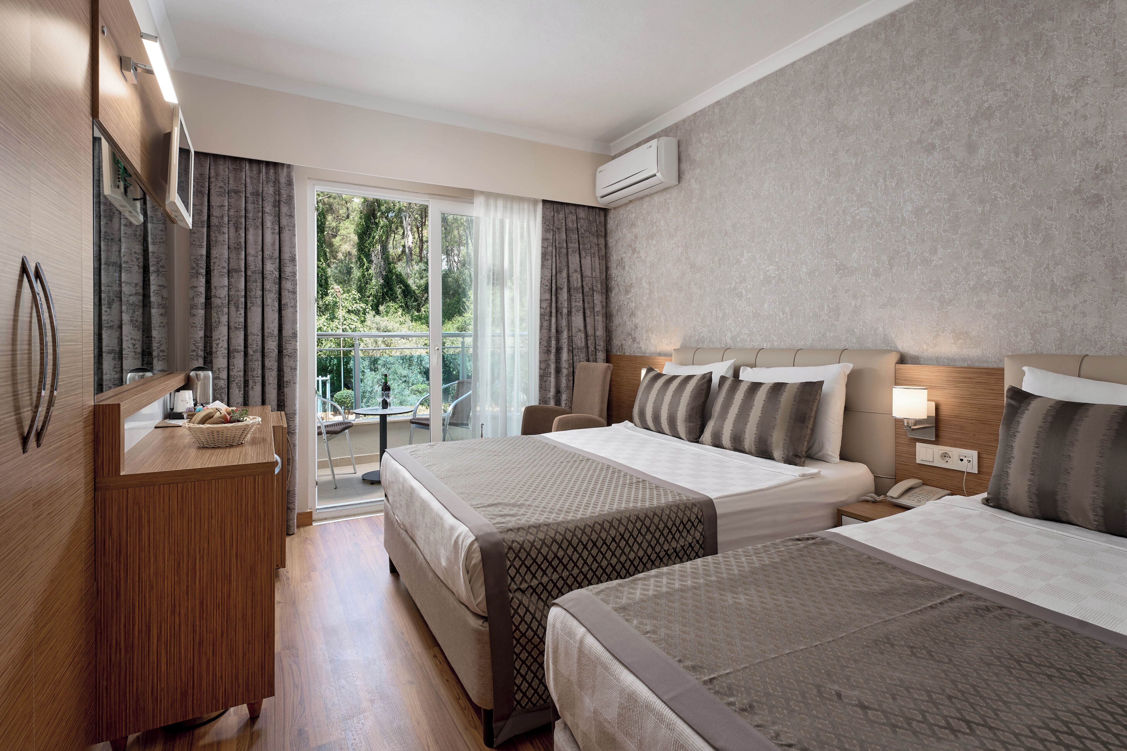 Quarto de hotel com duas camas, uma cômoda de madeira, uma escrivaninha e varanda com vista para área verde.