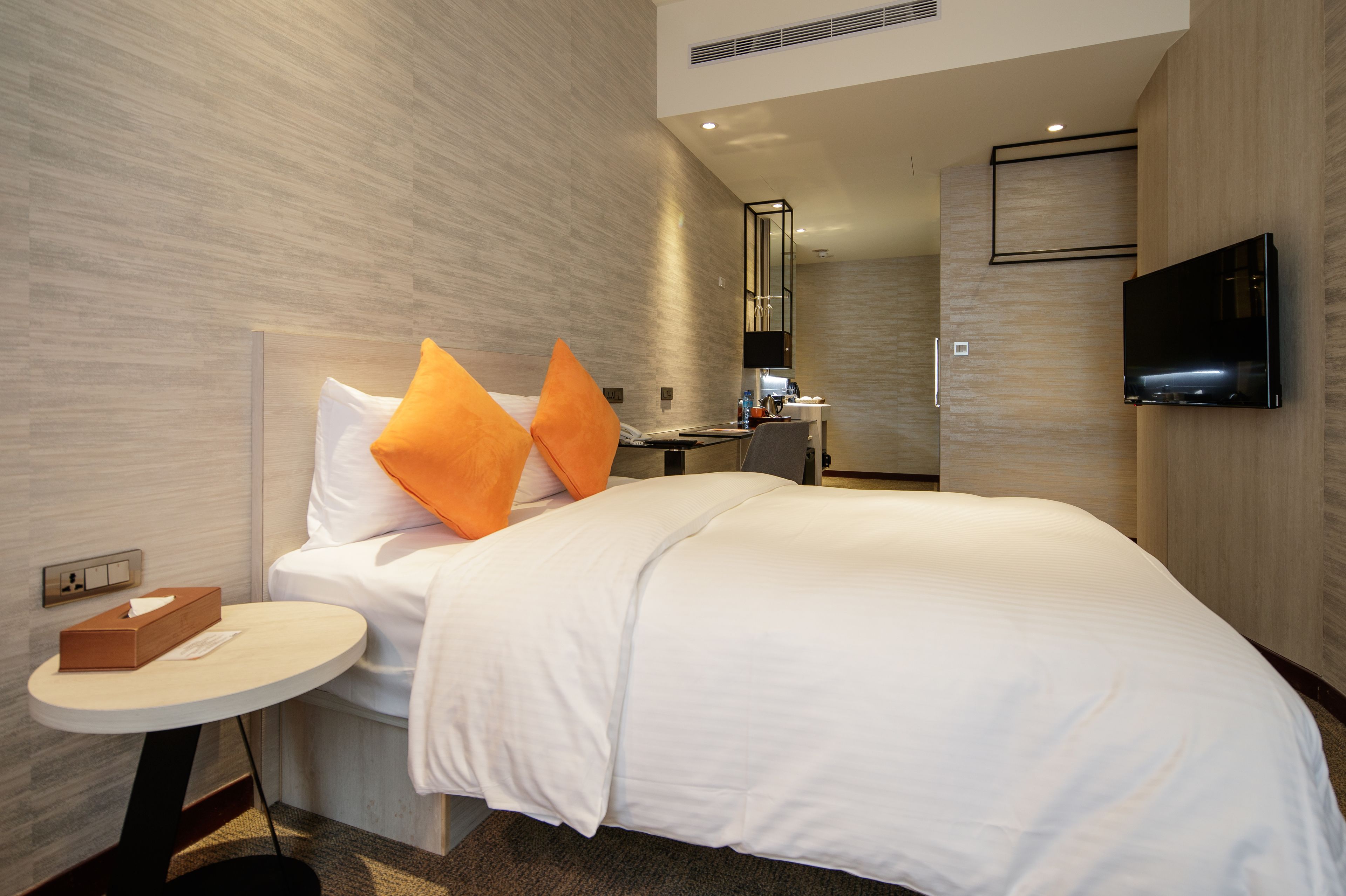 Quarto de hotel moderno com uma cama bem arrumada, uma pequena mesa de cabeceira com um porta-lenços, uma TV de tela plana na parede e uma área de cozinha compacta ao fundo.