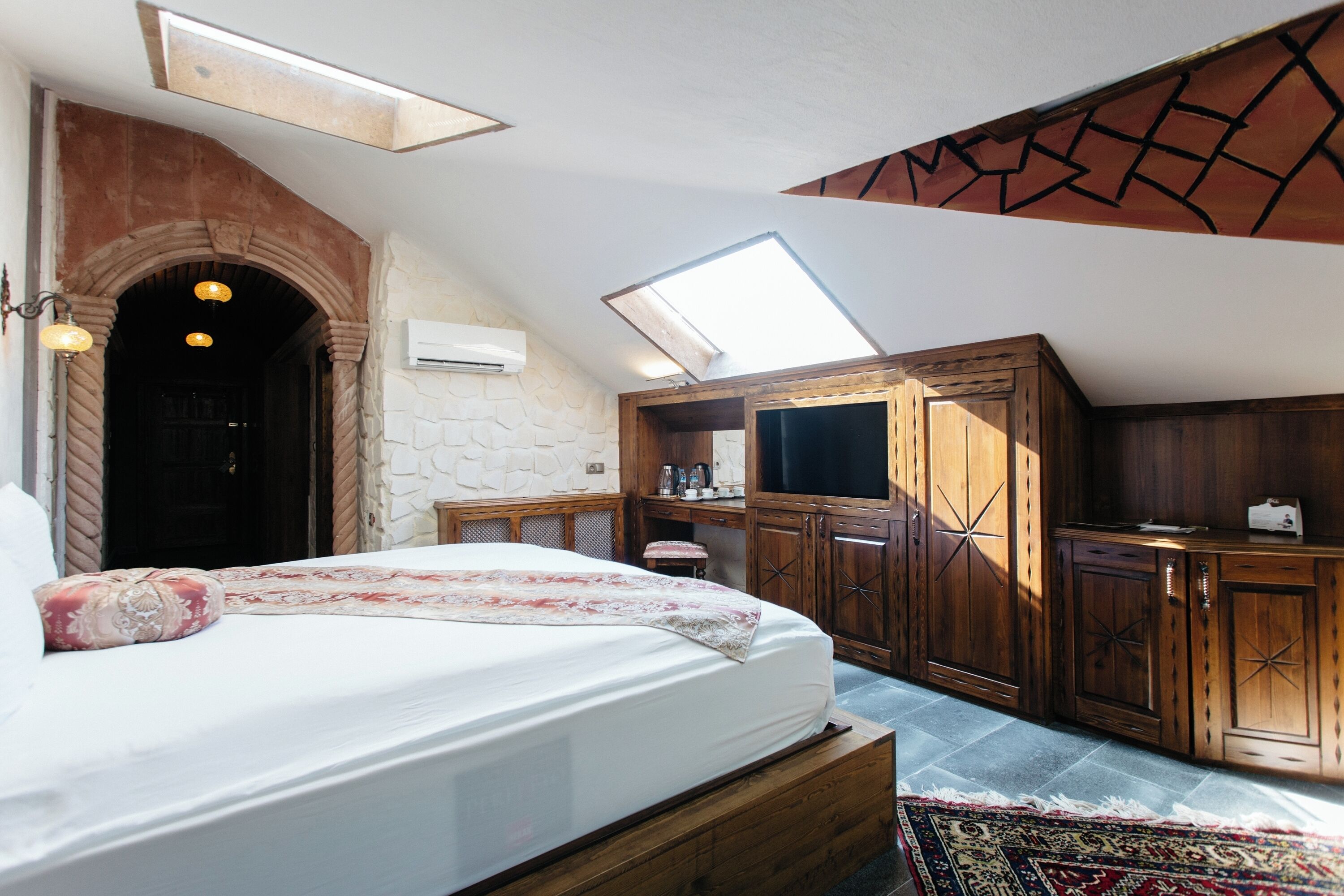 Quarto com cama, claraboia, parede de pedra, armários de madeira e televisão.