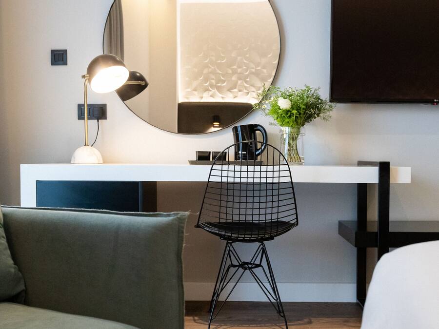 Quarto de hotel moderno com piso de madeira, sofá verde, cadeira de arame preta, escrivaninha com luminária branca, espelho redondo e TV de tela plana na parede.