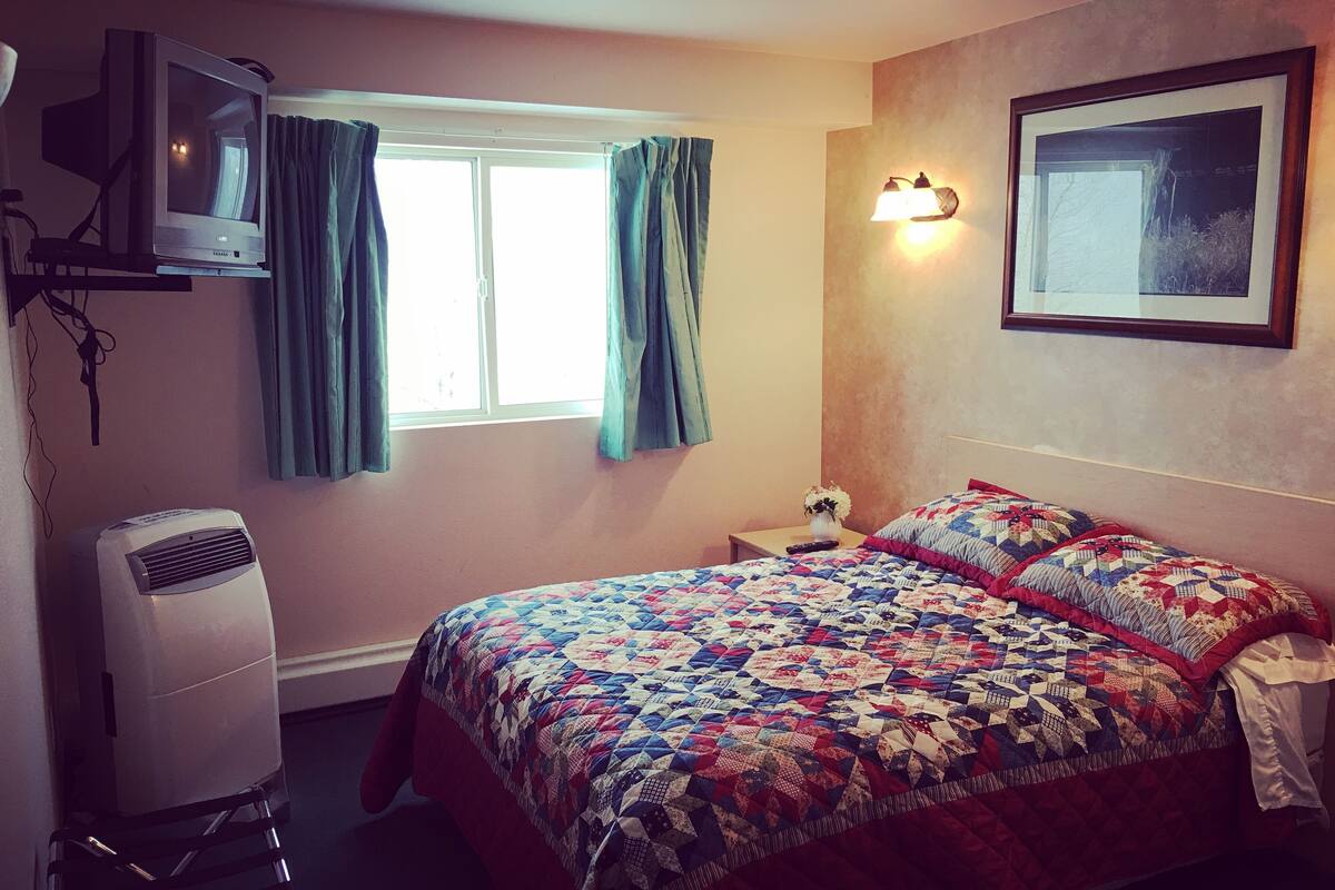 Quarto de hotel com cama, mesa de cabeceira com abajur, televisão na parede, janela com cortinas e porta-retrato fixado na parede.