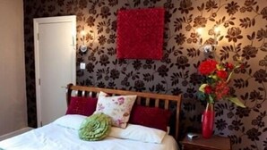 Um quarto com cama de madeira, roupa de cama branca, um travesseiro verde, travesseiros vermelhos e florais, um vaso vermelho com flores e uma parede com papel de parede floral.