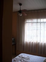 Quarto com cama, ventilador de teto, janela com cortinas leves e uma pequena mesa com televisão.
