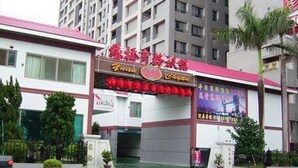 Entrada de hotel com uma placa vermelha e branca, um grande outdoor e uma palmeira ao primeiro plano.
