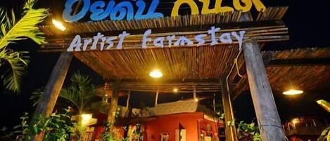 Um edifício rústico com uma placa que diz "Artist farmstay" e outra placa com escrita tailandesa.