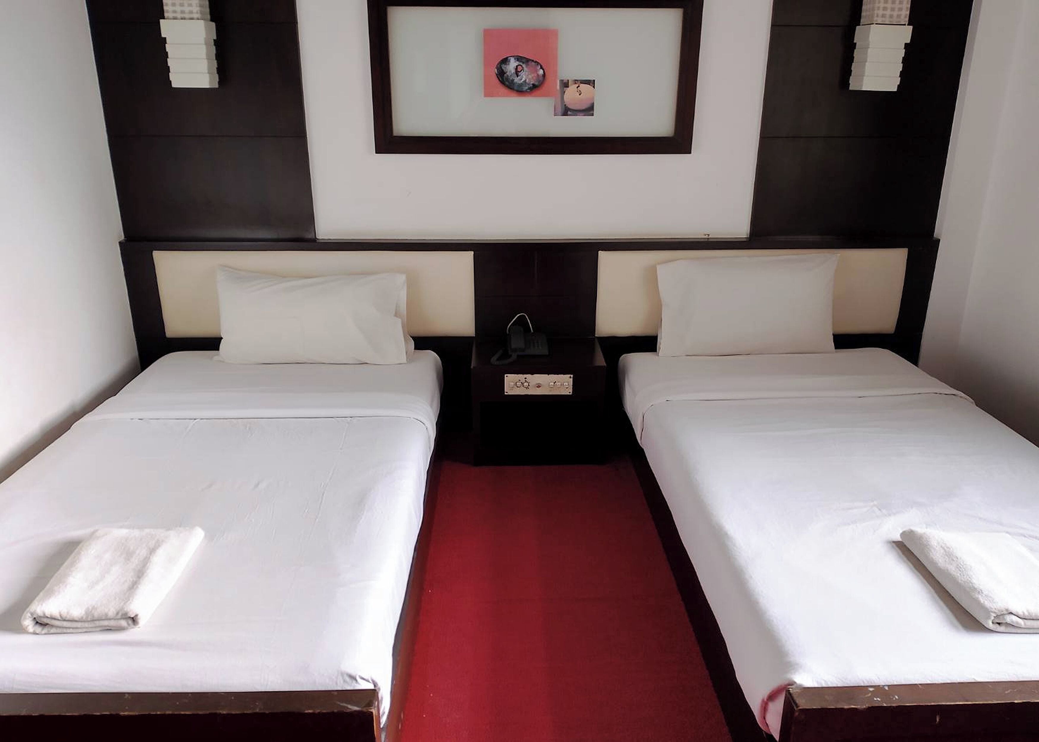Quarto de hotel com duas camas de solteiro, cada uma com roupa de cama branca e um tapete vermelho. Há uma pequena mesa com um telefone e uma tomada elétrica entre as camas.