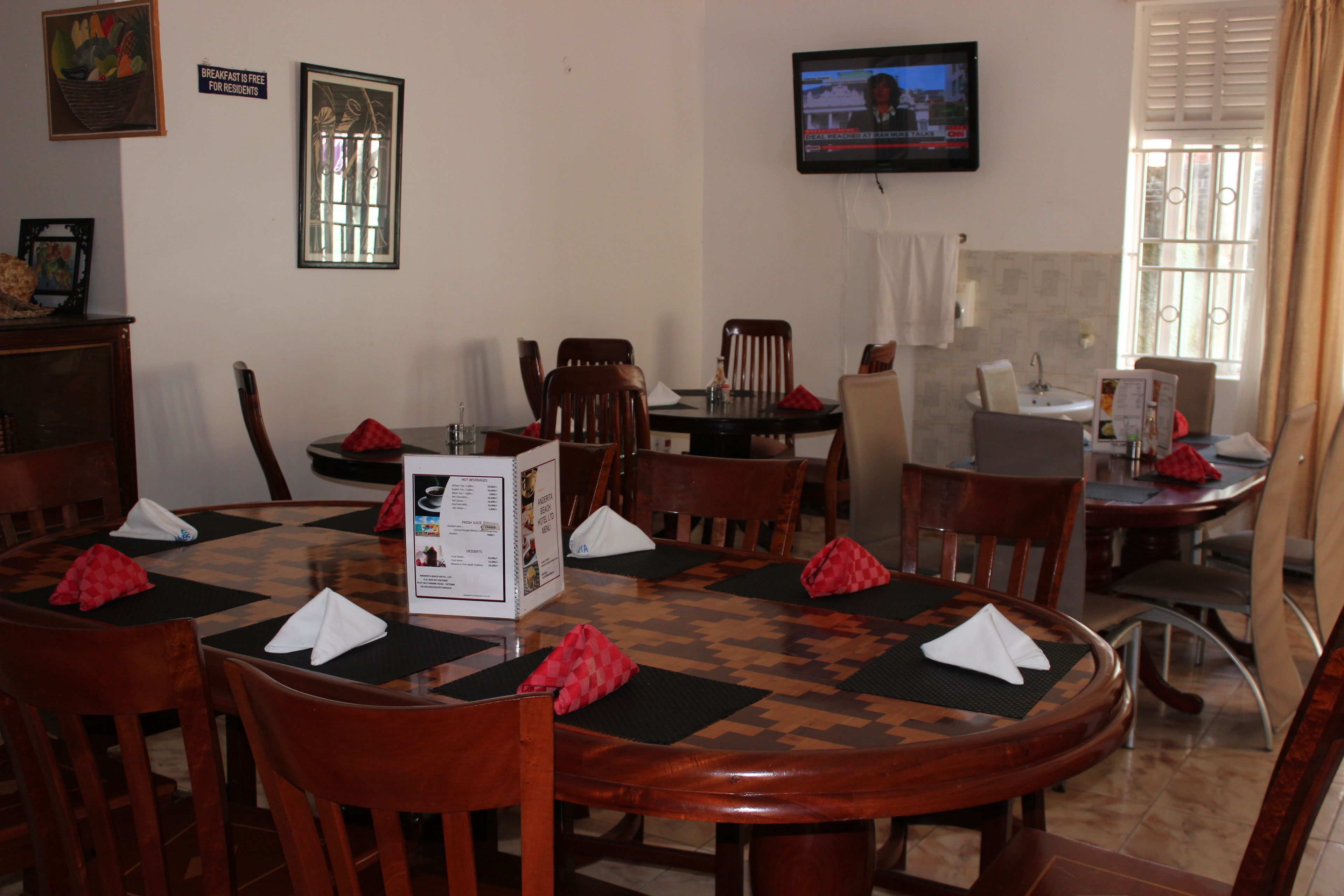 Uma área de refeições com mesa e cadeiras de madeira, um suporte para cardápio e uma televisão na parede.