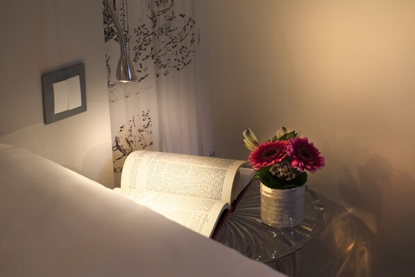 Cama bem arrumada com edredom branco e um vaso de vidro transparente com flores sobre uma mesa de cabeceira.