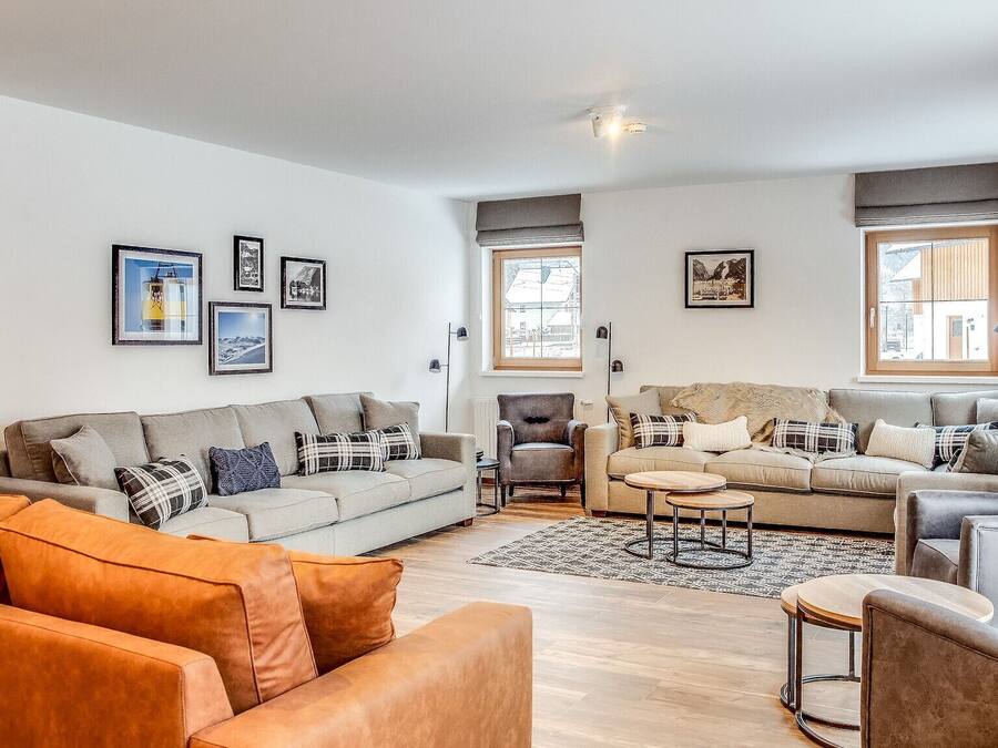 Uma sala de estar espaçosa com um amplo sofá, um sofá menor e uma mesa de centro. Há quadros nas paredes e uma janela que ilumina naturalmente o espaço.
