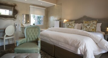 Quarto com uma cama grande, uma poltrona verde, uma toucador com espelho e janela com vista para árvores.