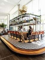 Uma área de bar moderna com balcão curvo, diversas opções de assento e uma escultura decorativa de galo dourado.