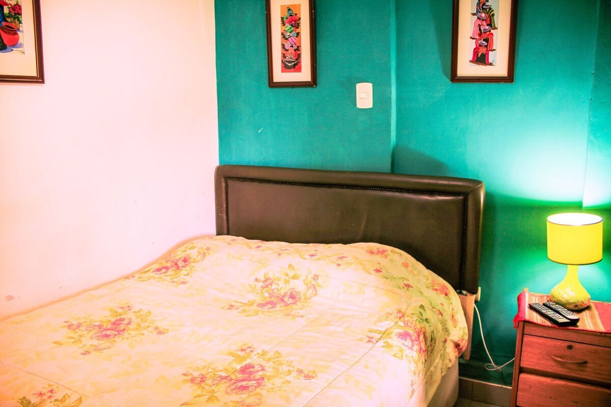 Uma cama com colcha florida, uma mesa de cabeceira com abajur e duas fotos emolduradas em paredes verde-turquesa.