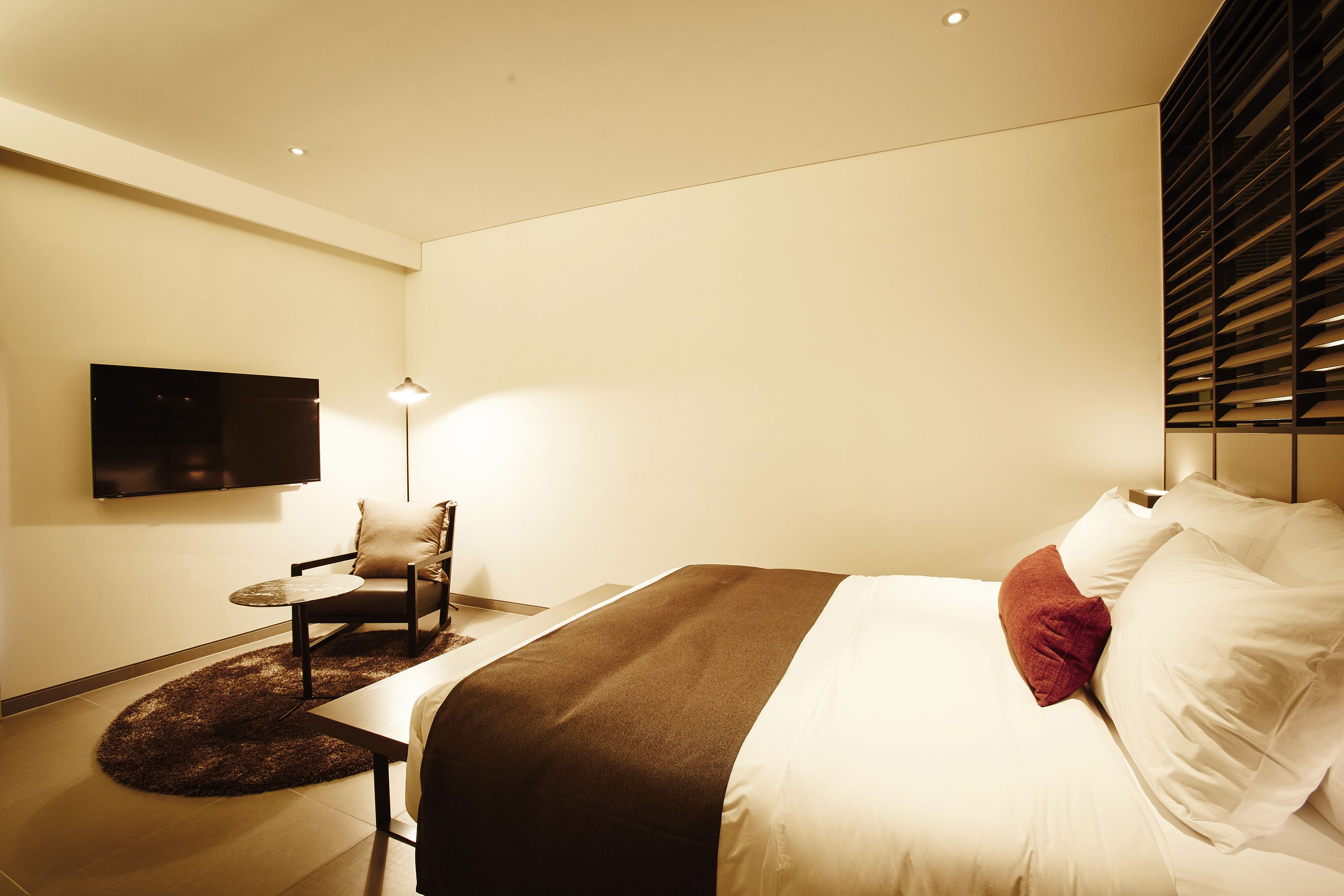 Quarto de hotel moderno com cama, uma cadeira, uma mesinha e uma TV de tela plana fixada na parede.