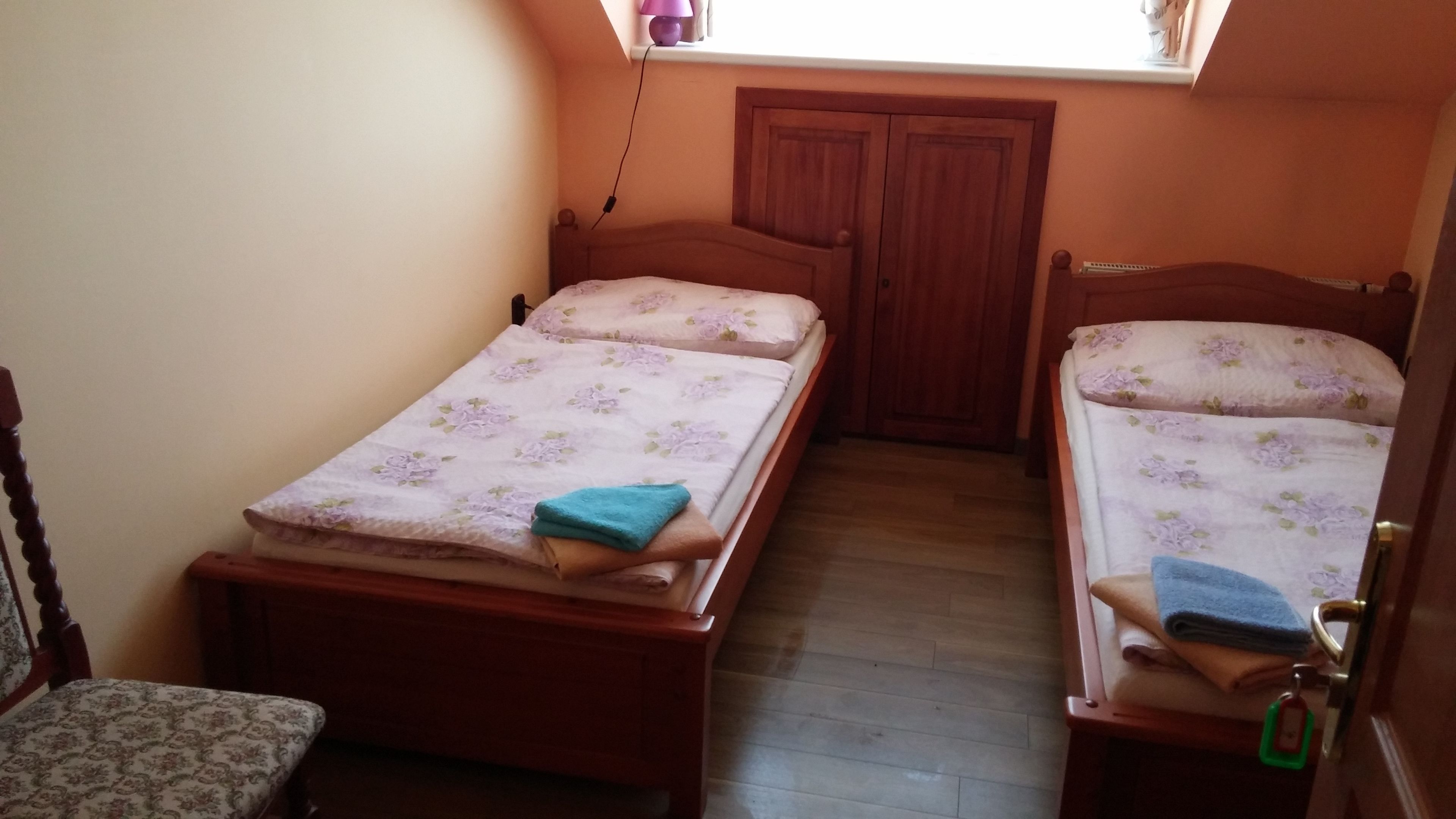 Quarto pequeno com duas camas de solteiro, um guarda-roupa de madeira, uma cadeira e uma janela com luminária.